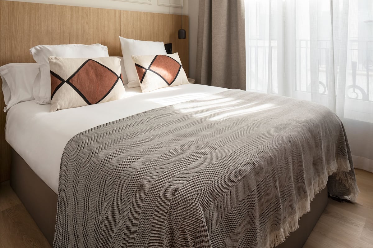 Cama bem arrumada com colcha estampada, cabeceira de madeira e uma janela grande com cortinas translúcidas.