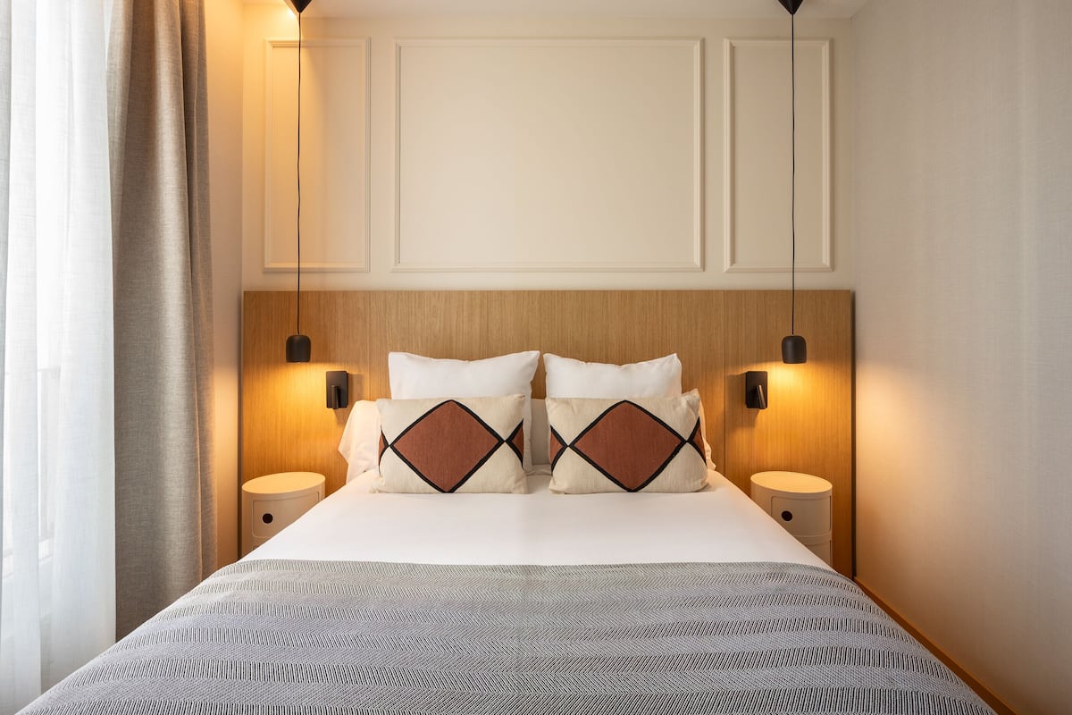 Quarto de hotel com cama, duas mesas de cabeceira, painel de cabeceira e luminárias fixadas na parede.