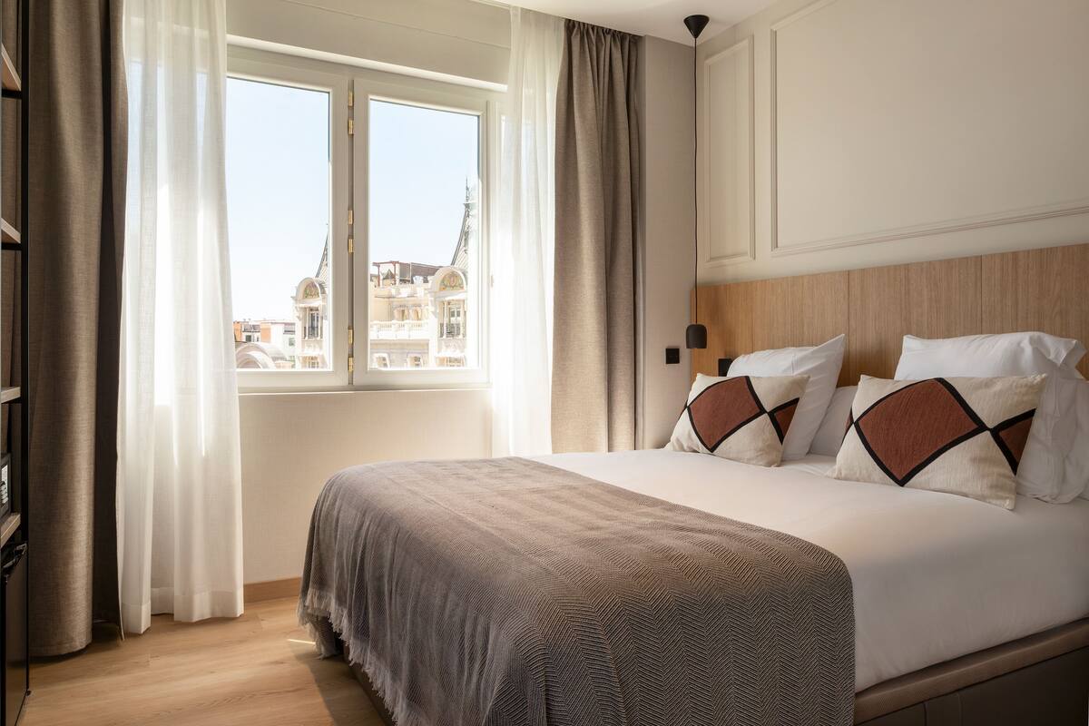 Quarto de hotel com uma cama grande, uma janela com cortinas, um painel de cabeceira de madeira e vista para edifícios.