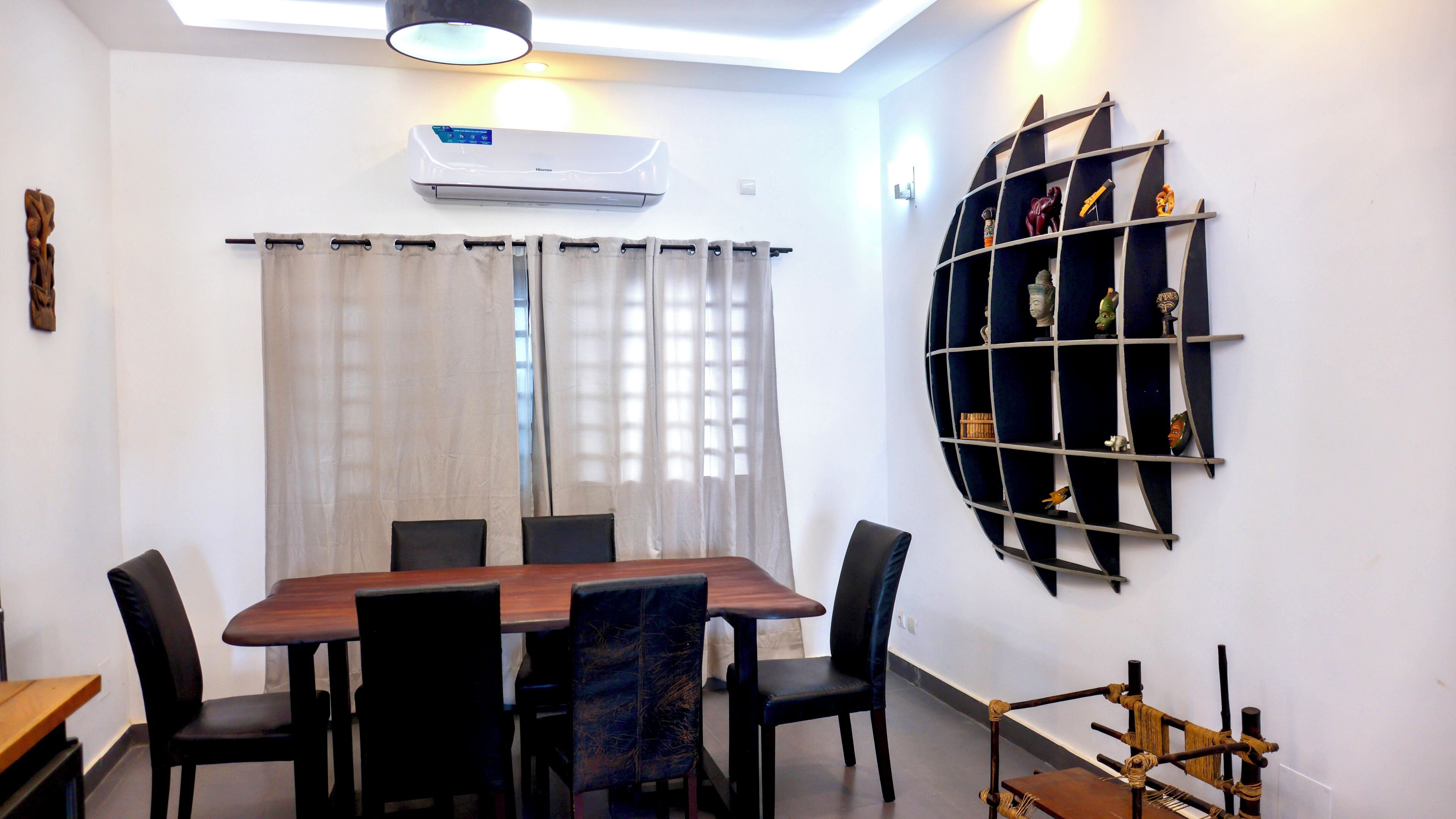 Uma sala de jantar com uma mesa de madeira, cadeiras pretas, uma prateleira fixada na parede com objetos decorativos e um aparelho de ar condicionado.