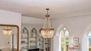 Quarto com televisão em um armário branco, lustre, prateleiras com objetos decorativos e janela com venezianas.