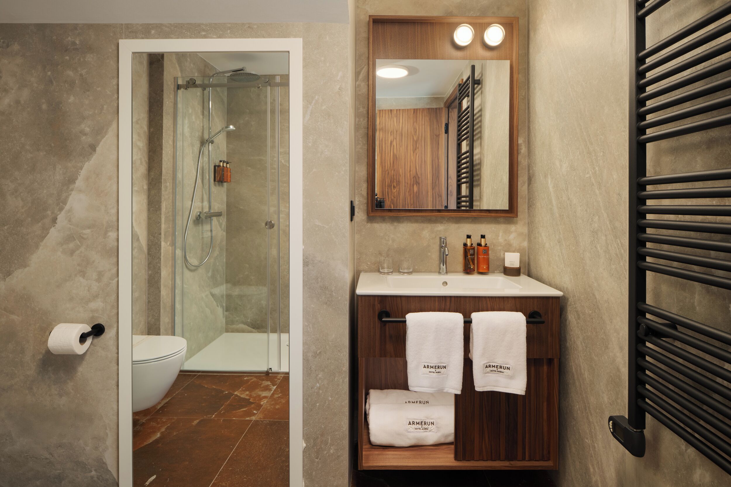 Um banheiro com box de vidro, vaso sanitário, pia com toucador de madeira e porta-toalhas com toalhas.
