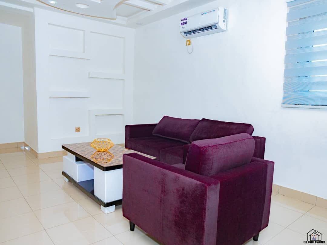 Uma sala de estar com um sofá roxo escuro, uma pequena mesa de centro de madeira e uma parede branca com teto rebaixado.
