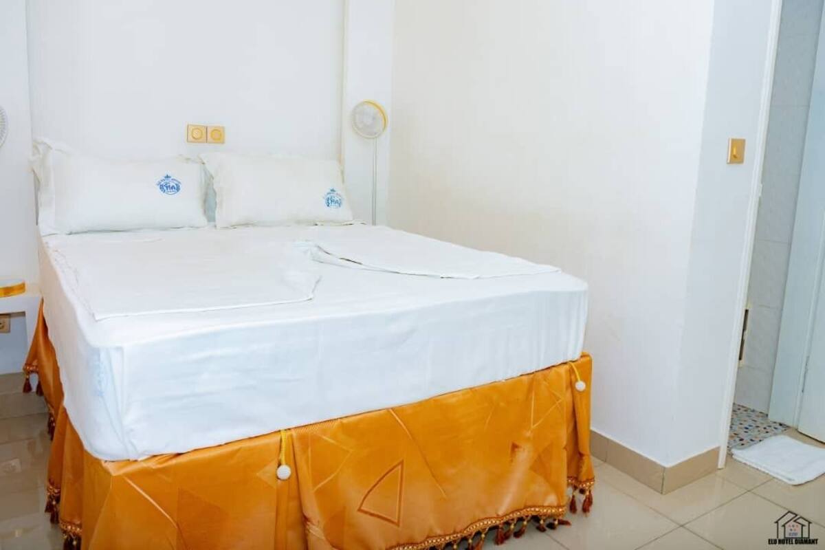Uma cama de solteiro com colchão branco e saia de cama laranja em um quarto com paredes brancas e piso de azulejos.