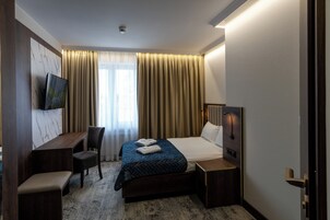 Quarto de hotel com cama, escrivaninha, cadeira, televisão e mesa de cabeceira.