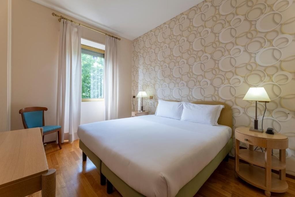 Quarto de hotel com uma cama grande, duas mesas de cabeceira com abajures, uma cadeira de madeira e uma janela com cortinas.