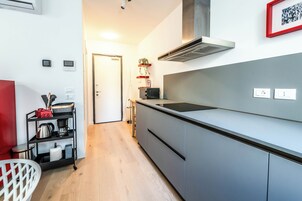 Cozinha moderna com armários cinza, bancada preta e exaustor de aço inoxidável. Há uma pequena prateleira com uma planta e um quadro com moldura vermelha na parede.