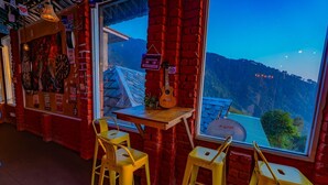 Um espaço de bar acolhedor com paredes de tijolo, uma mesa de madeira e banquetas amarelas. Há uma guitarra sobre a mesa e uma vista de montanha através de uma grande janela.