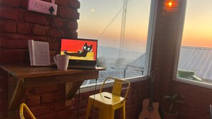 Um quarto com parede de tijolos, um laptop exibindo um desenho animado, uma guitarra e uma janela com vista para painéis solares.