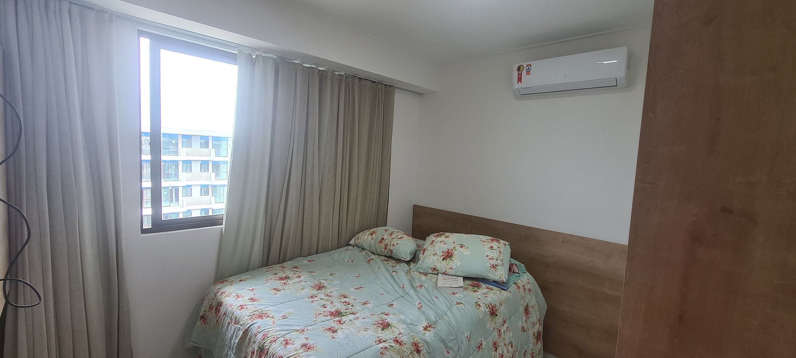 Quarto com cama, cortinas, aparelho de ar condicionado e janela com vista para edifícios.