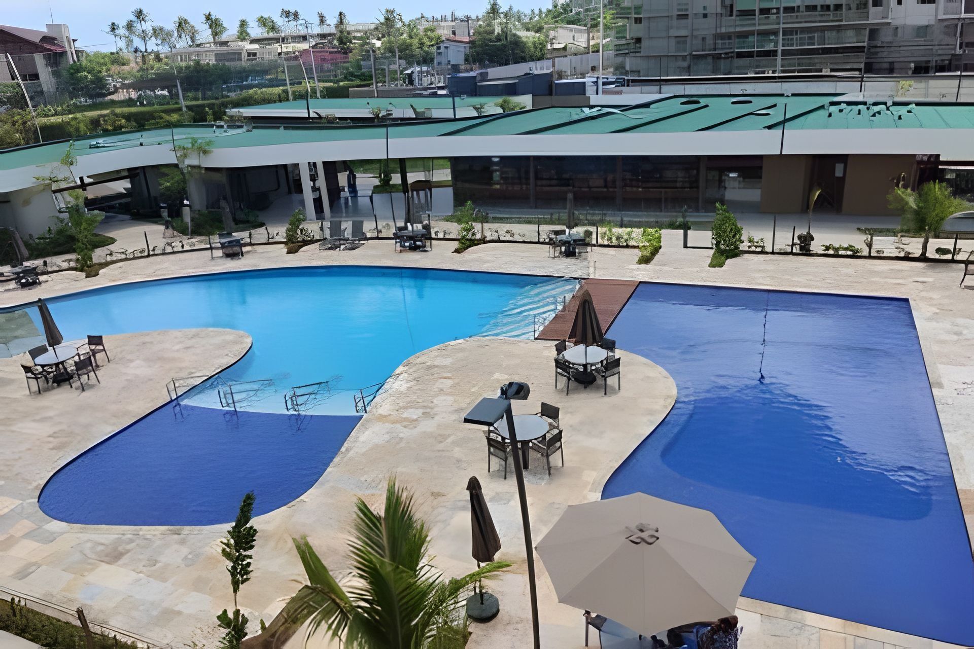 Piscina com duas áreas distintas, rodeada por espreguiçadeiras e guarda-sóis, com vista para um edifício ao fundo.