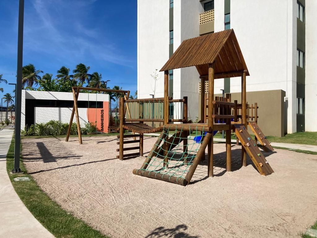 Parquinho infantil com estrutura de escalada e escorregador, com um prédio de fachada branca e uma palmeira ao fundo.