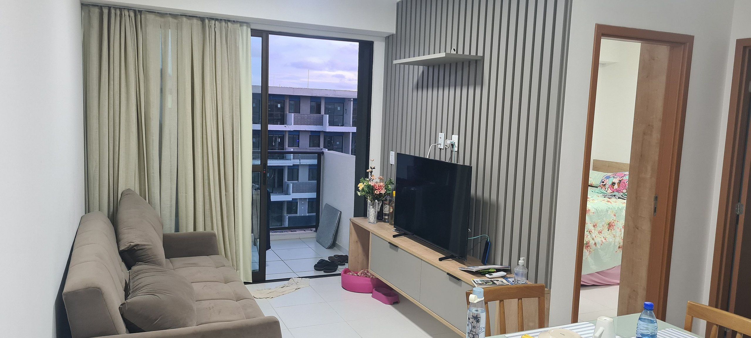 Sala de estar com sofá, televisão de tela plana, mesa de jantar e vista para um edifício através de uma porta de vidro.