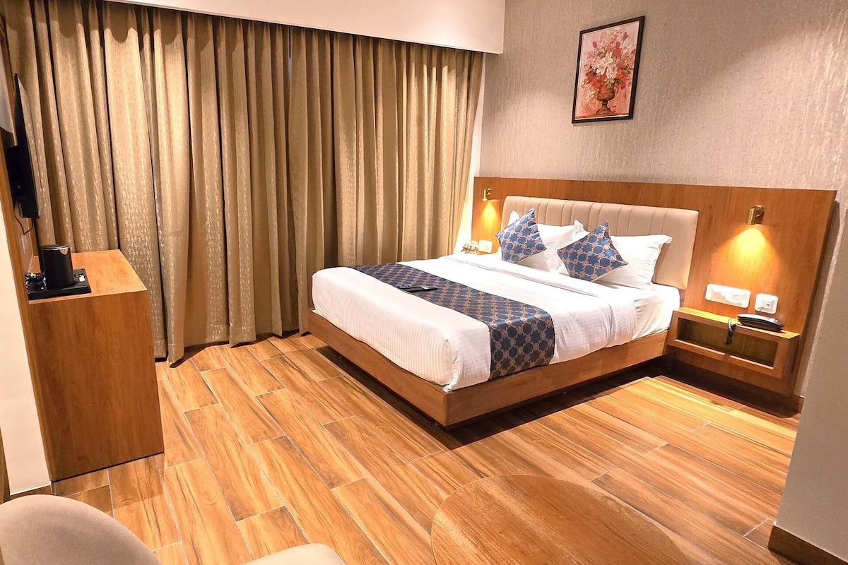 Quarto de hotel com cama de madeira, escrivaninha, cadeira, mesa de cabeceira e televisão fixada na parede.