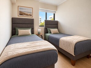 Quarto com duas camas, uma mesinha de cabeceira, abajur e janela com vista para edifícios.