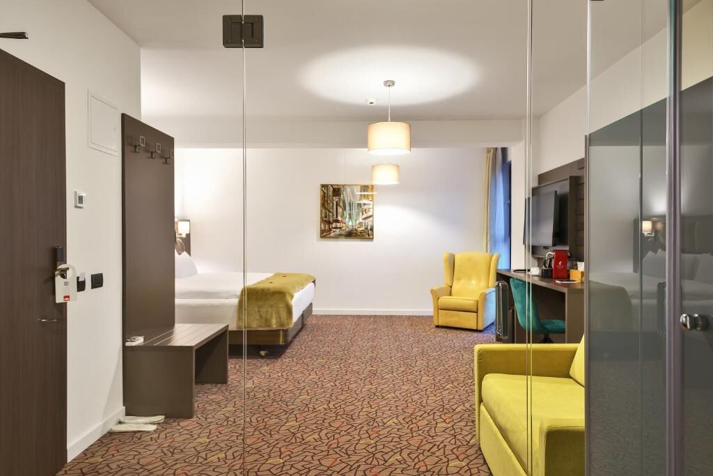 Quarto de hotel com cama, escrivaninha, cadeira, televisão e um quadro na parede.