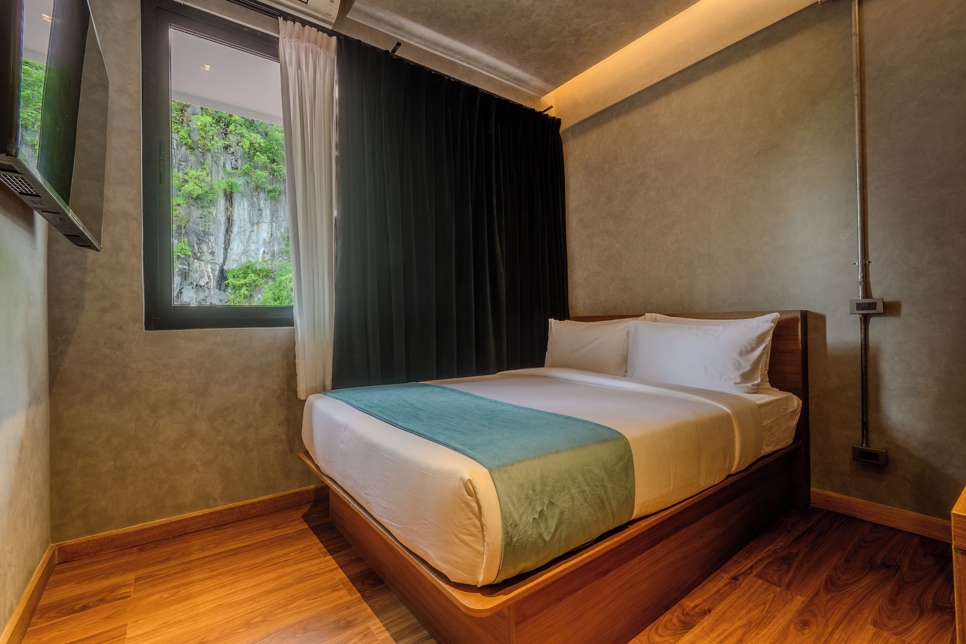 Uma cama bem arrumada com um manta verde, uma janela com cortinas, uma televisão de tela plana e um luminário fixado na parede.