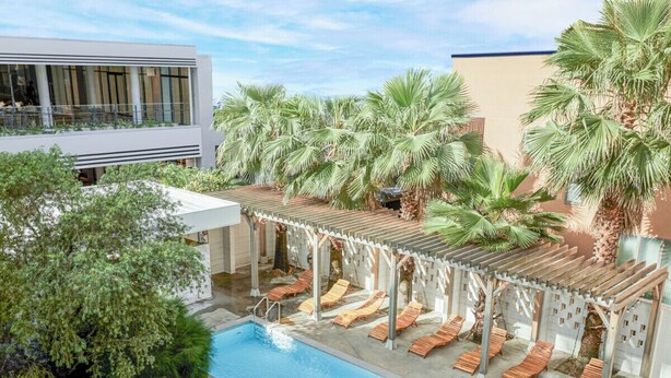 Piscina no terraço com espreguiçadeiras e guarda-sóis, cercada por palmeiras e um edifício com grandes janelas.