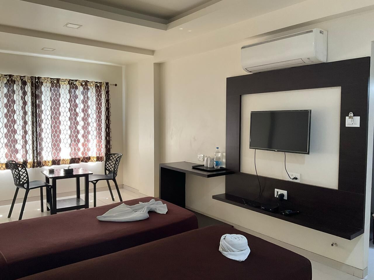 Quarto de hotel com televisão, uma pequena mesa com cadeiras, um sofá e uma janela com cortinas.