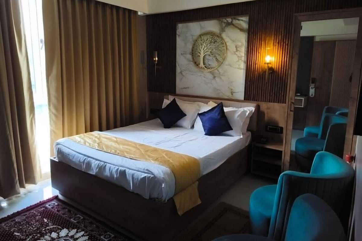 Quarto de hotel com cama, ventilador de teto, obra de arte na parede, área de estar com cadeiras e uma janela grande com cortinas.