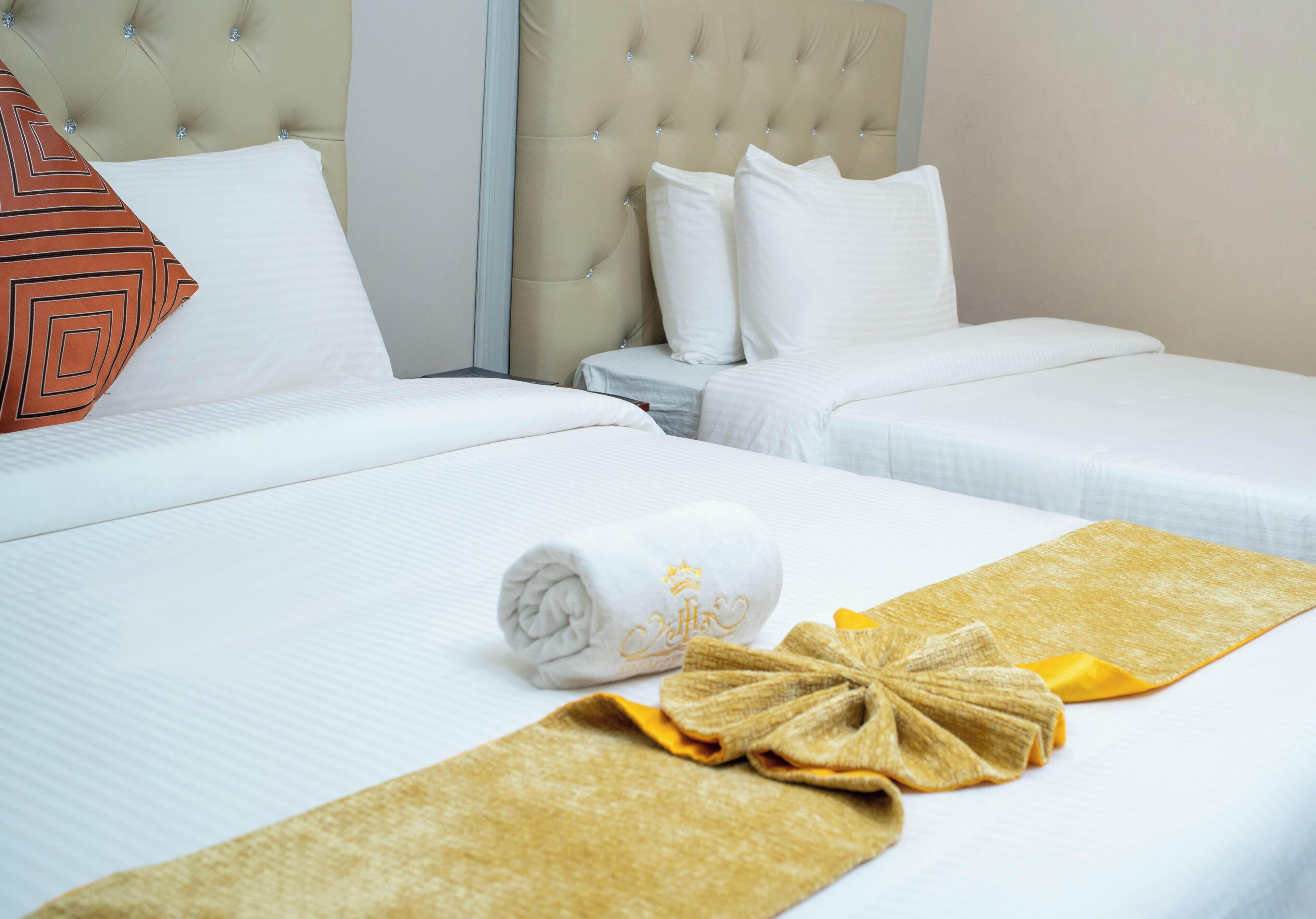 Quarto de hotel com duas camas, cabeceira estofada, roupa de cama branca e toalha enrolada com emblema dourado.