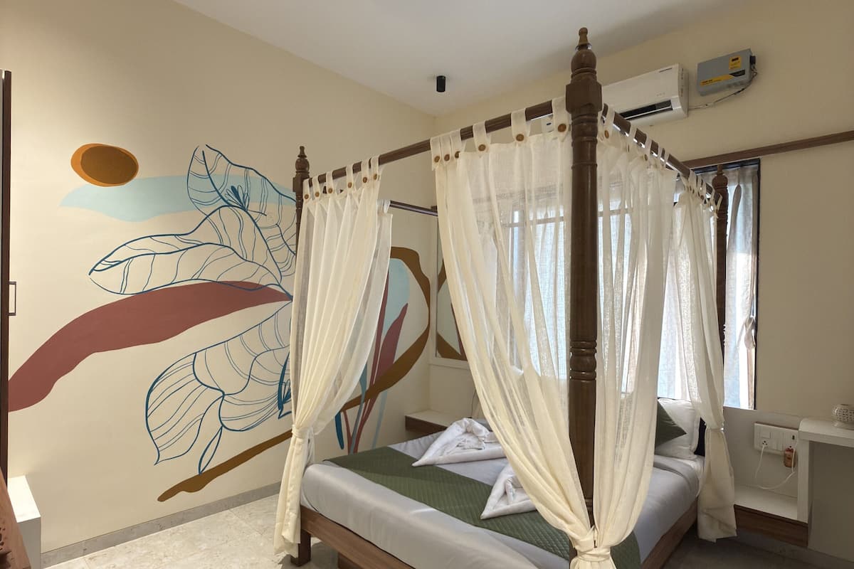 Uma cama com dossel e colunas, estrutura de madeira e cortinas brancas, um ventilador de teto e um aparelho de ar condicionado na parede.
