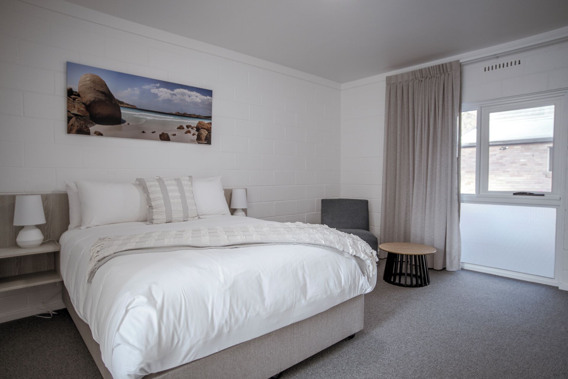 Um quarto com cama, luminárias de cabeceira, uma cadeira, uma mesinha, uma janela com cortinas e uma pintura de uma paisagem praiana na parede.