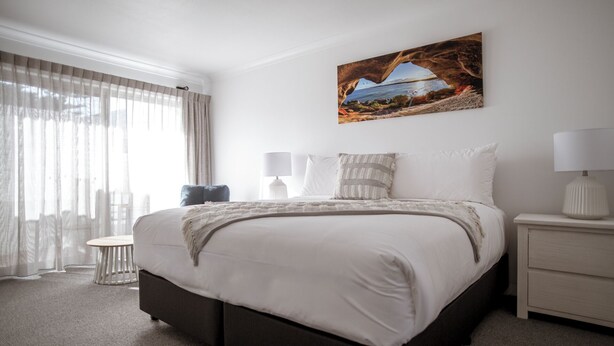 Quarto de hotel com cama, mesa de cabeceira, luminária, cadeira e uma janela grande com cortinas.