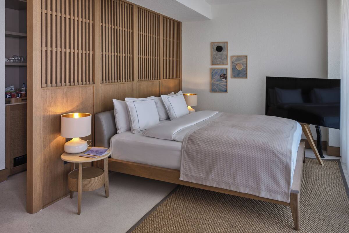 Quarto de hotel com cama, luminárias de cabeceira, televisão e divisória de madeira.