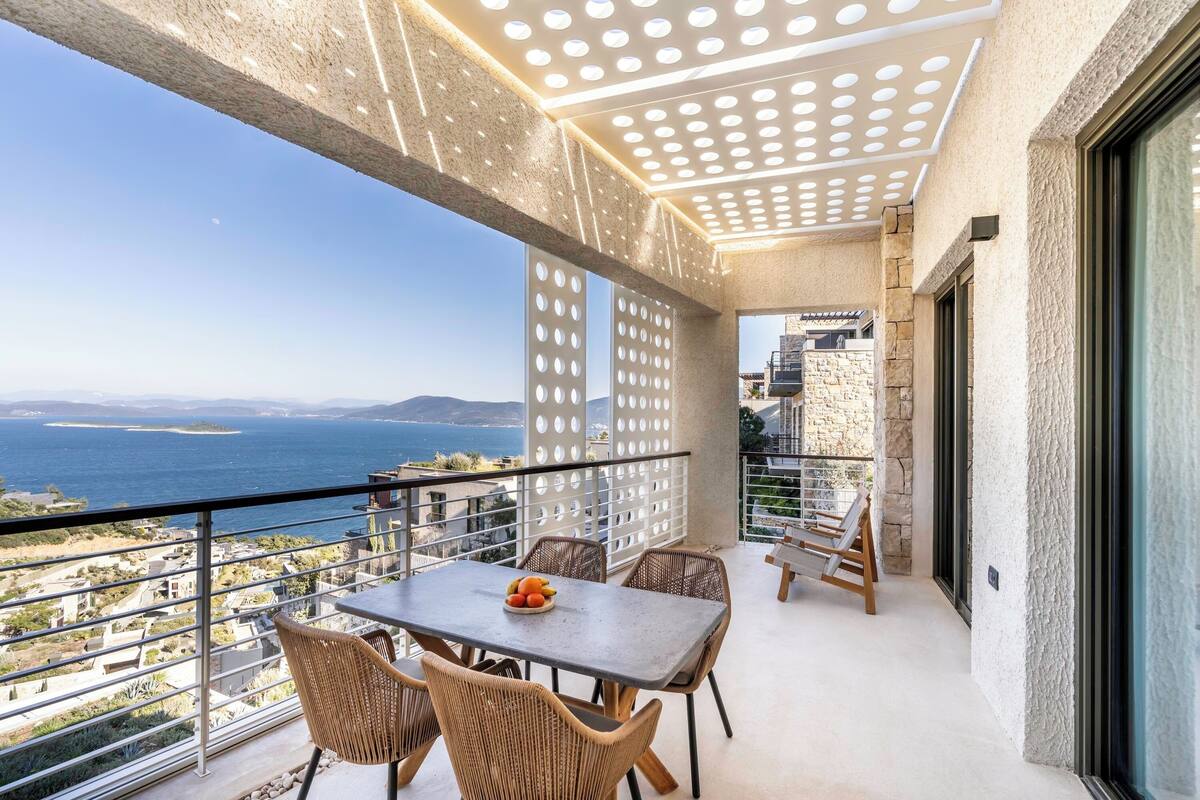 Uma varanda com mesa e cadeiras, vista para o mar e um horizonte urbano ao fundo.