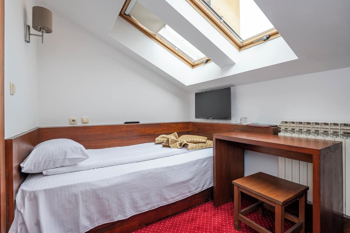 Quarto compacto com cama, escrivaninha, televisão e claraboia.