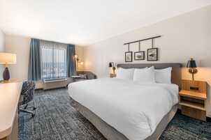 Quarto de hotel com uma cama grande, uma escrivaninha, uma cadeira, uma luminária de mesa, uma mesa de cabeceira, uma luminária de parede e uma janela com cortinas.