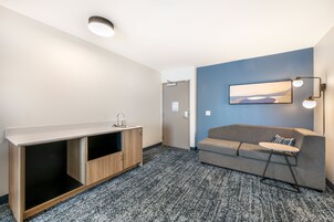Quarto de hotel com cozinha compacta, sofá, mesa pequena e luminária fixada na parede.