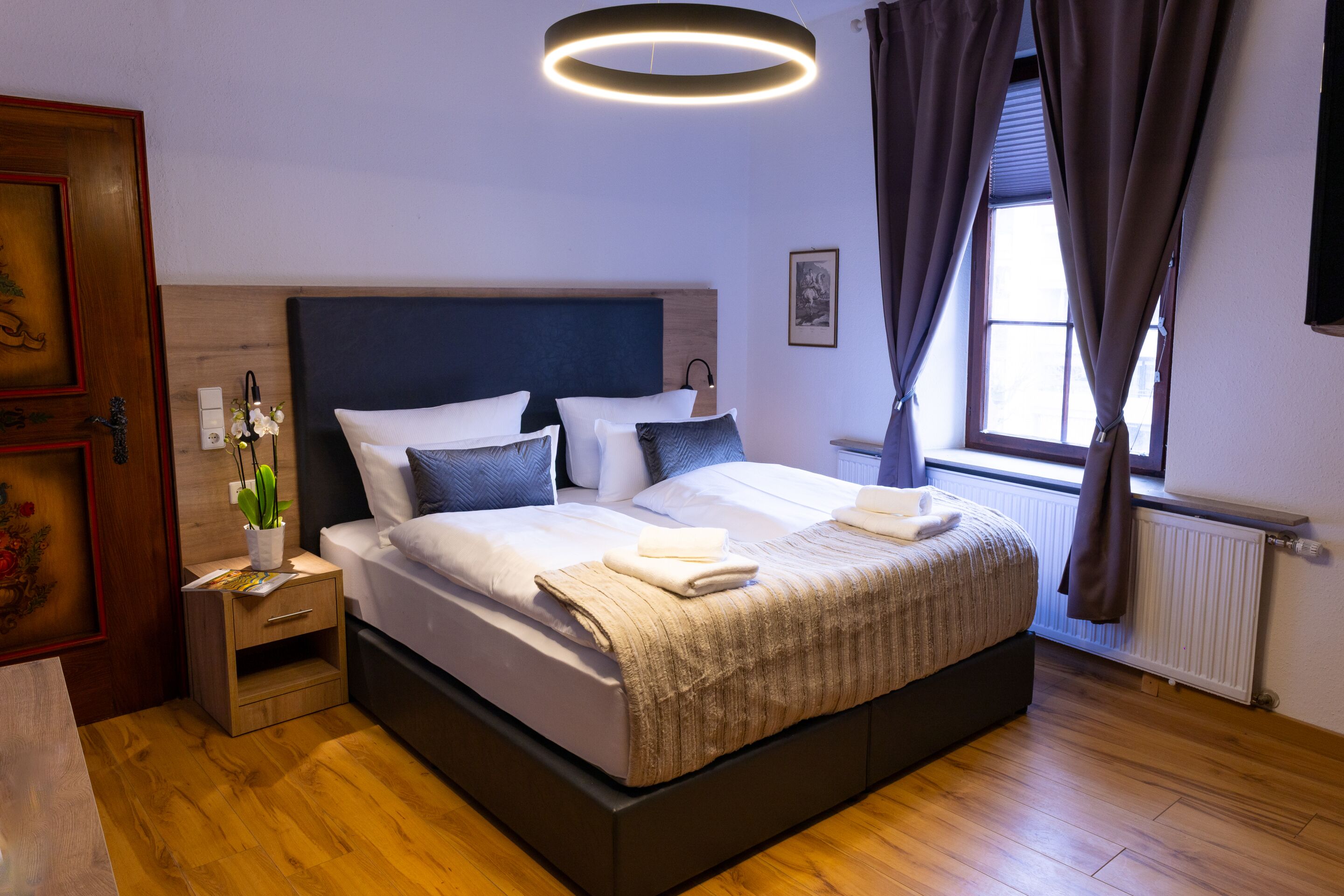 Quarto moderno com uma cama grande, mesas de cabeceira, um criado-mudo, uma luminária de parede e uma janela com cortinas.