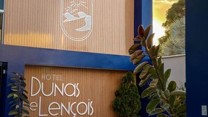 Exterior do Hotel DUNAS e Lencois com fachada de madeira, um logotipo com um sol e ondas, e uma placa com o nome do hotel.