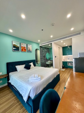 Quarto de hotel moderno com uma cama grande, uma escrivaninha, uma cozinha compacta e um banheiro com box de vidro.