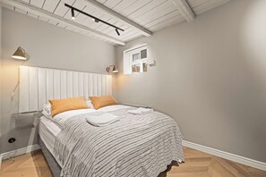 Quarto com cabeceira branca, colcha listrada e duas almofadas. Há uma janela com vista para o exterior, uma mesa de cabeceira com abajur e piso de madeira.