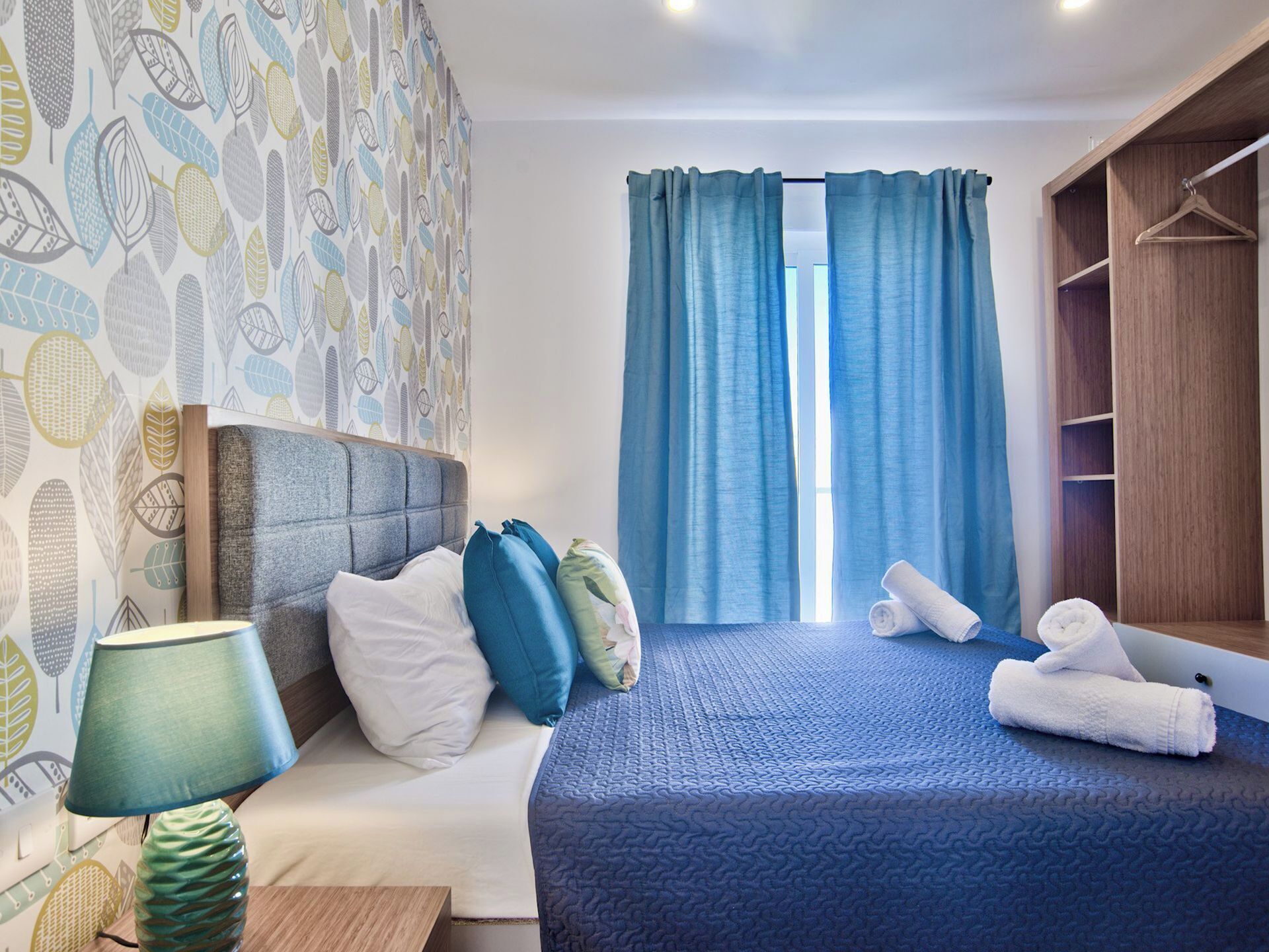 Quarto com cama, almofadas, abajur, guarda-roupa e cortinas.