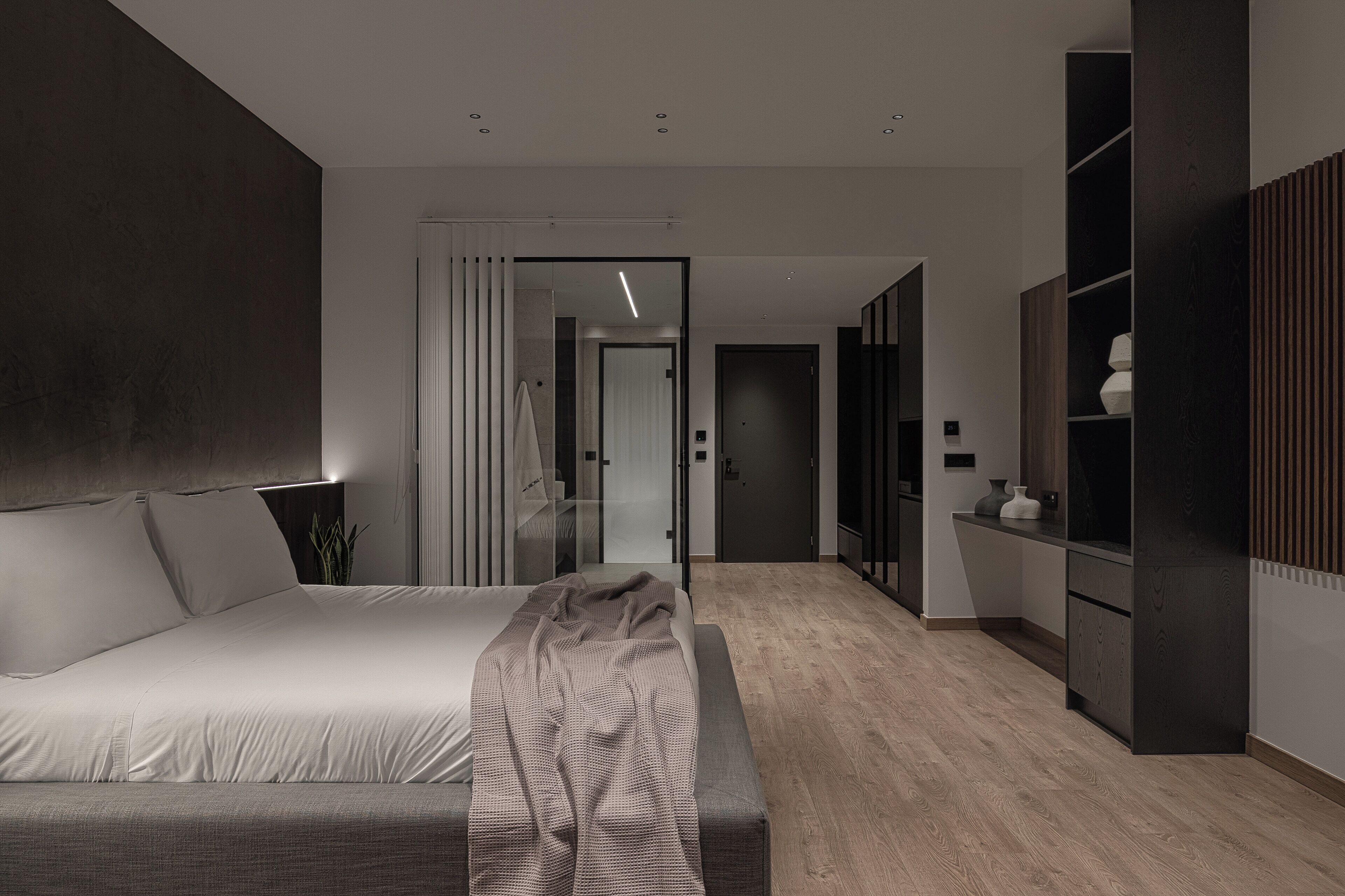 Quarto moderno com cama grande, mesa de cabeceira, guarda-roupa e banheiro visível através de uma porta de vidro.