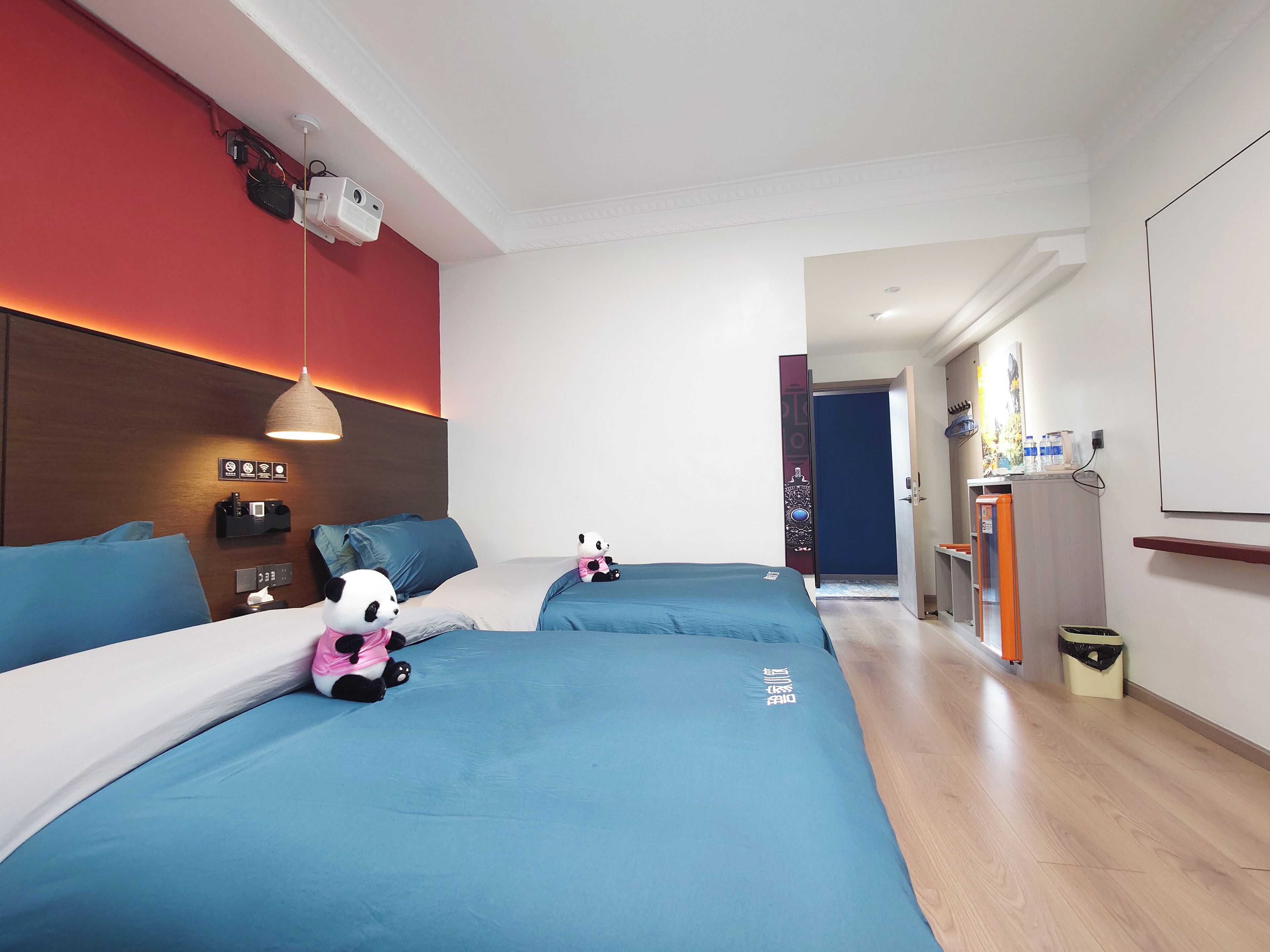 Quarto de hotel com uma cama, mesas de cabeceira, uma luminária, um projetor fixado na parede e uma porta para o banheiro.