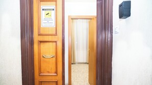 Uma porta de madeira com placa de segurança, corredor com piso ladrilhado e porta entreaberta dando acesso a outro cômodo.