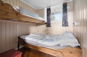 Uma cama beliche com cabeceira de madeira, uma janela com cortinas e uma mesinha.