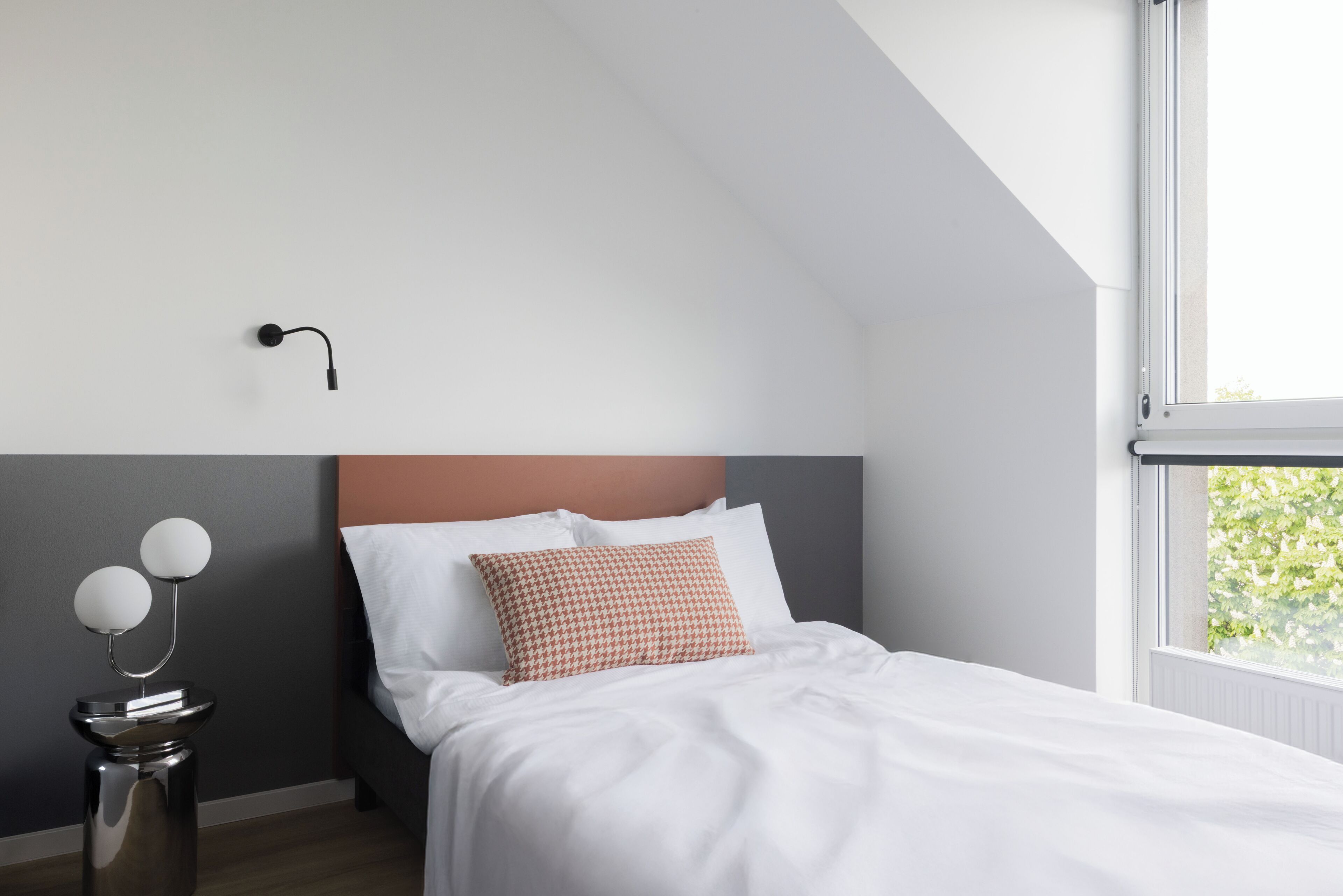 Quarto com cama, mesa de cabeceira, luminária na parede e janela.
