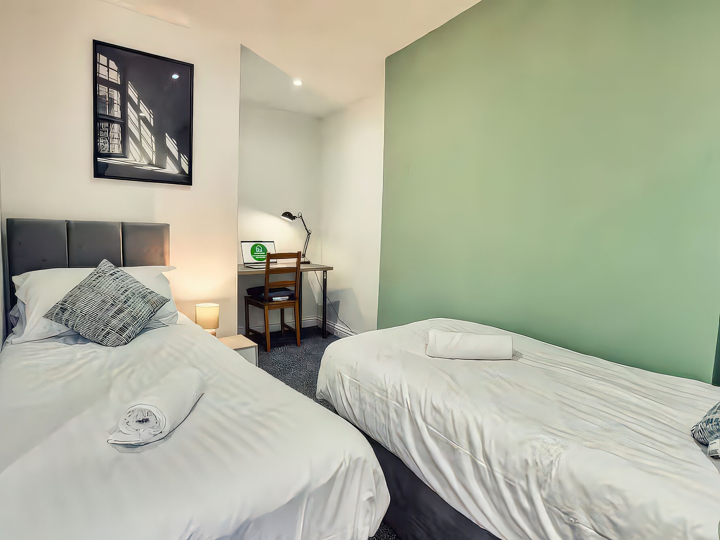 Quarto de hotel com duas camas, uma escrivaninha com um laptop, uma cadeira e um quadro na parede.
