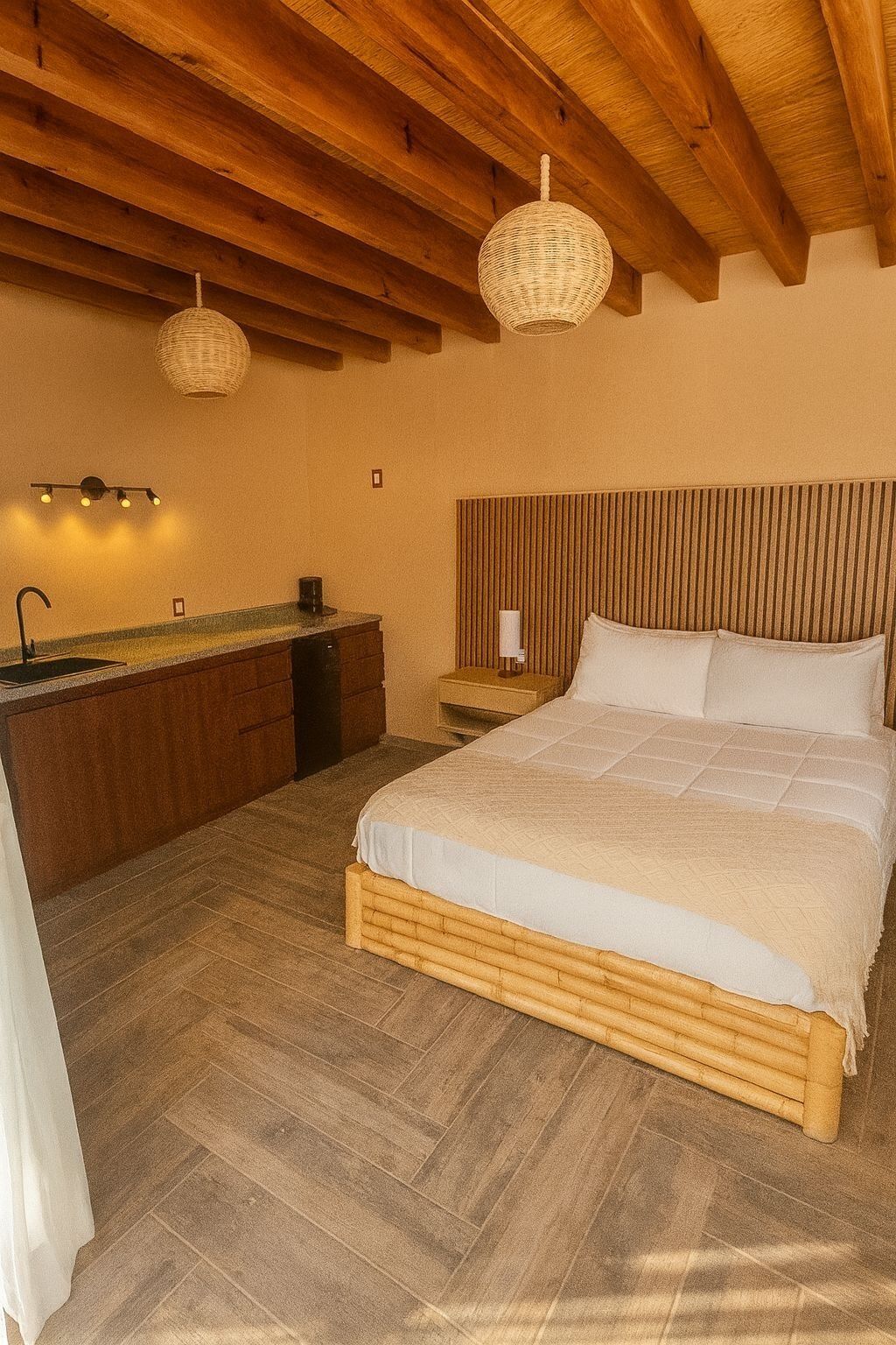 Quarto com cama de madeira, mesa de cabeceira, luminária pendente e vista para a cozinha.