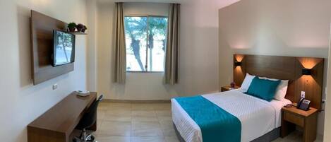 Quarto de hotel com cama, mesas de cabeceira, escrivaninha, cadeira, televisão na parede e janela com cortinas.