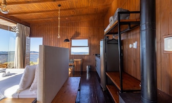 Interior de cabana com cama, uma mesinha, luminária pendente, janela com vista e um objeto cilíndrico preto.