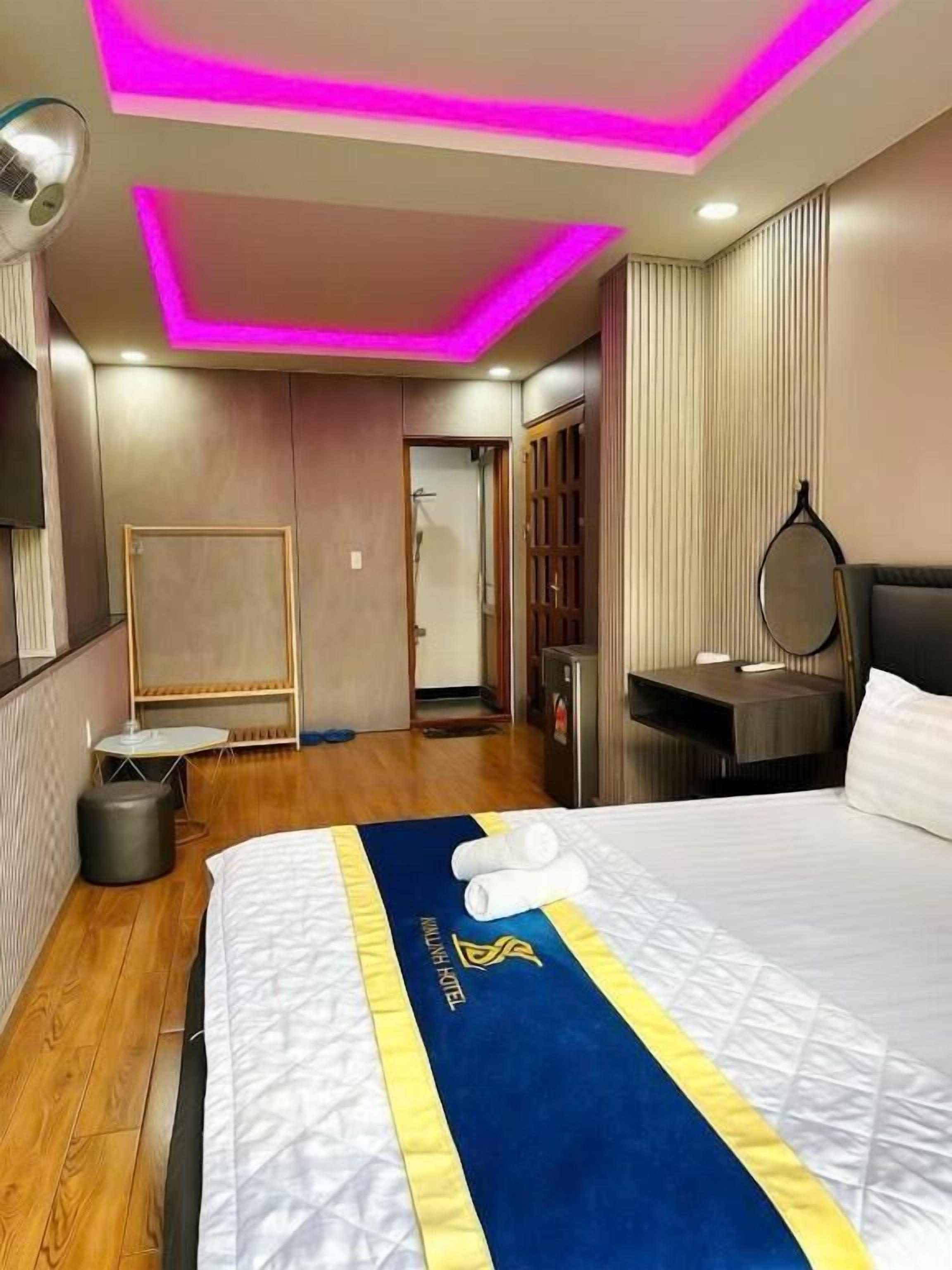Quarto de hotel com cama, mesa de cabeceira, espelho e iluminação rosa no teto.