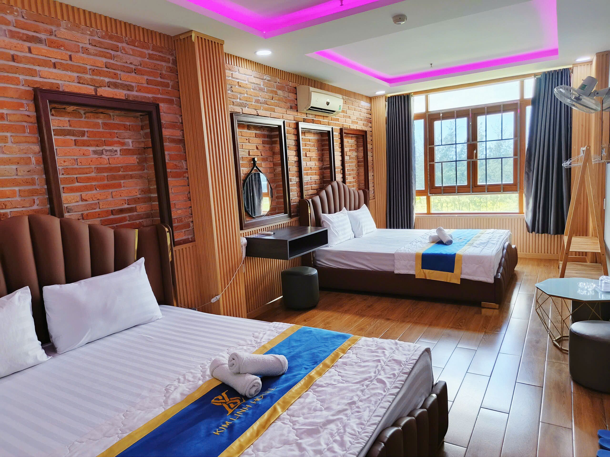 Quarto de hotel com duas camas, uma parede de destaque em madeira, uma janela grande com cortinas e iluminação rosa no teto.