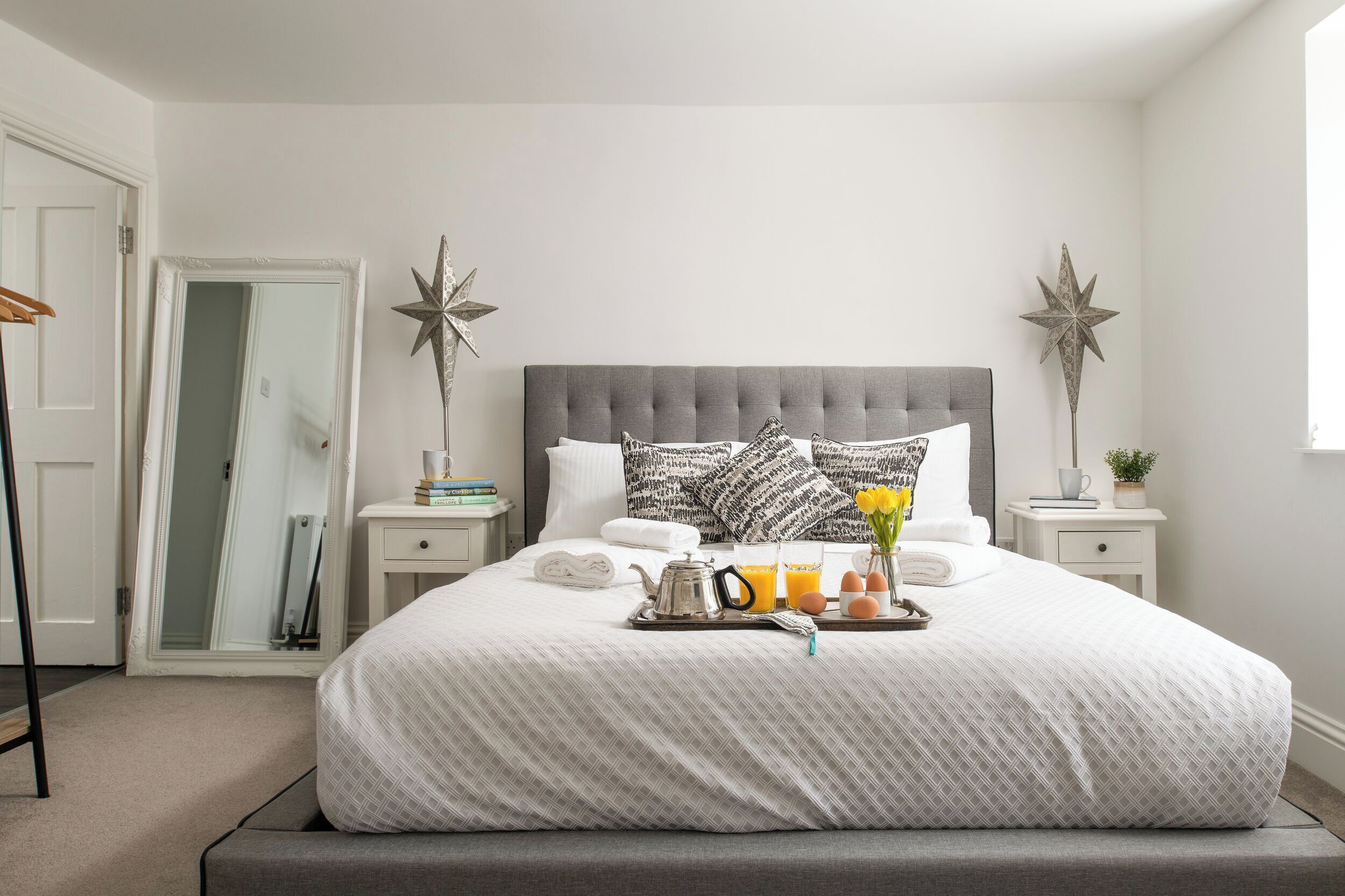Cama bem arrumada com cabeceira cinzenta, roupa de cama branca e almofadas decorativas. Há uma bandeja com bule, xícaras e um vaso de flores sobre a cama.